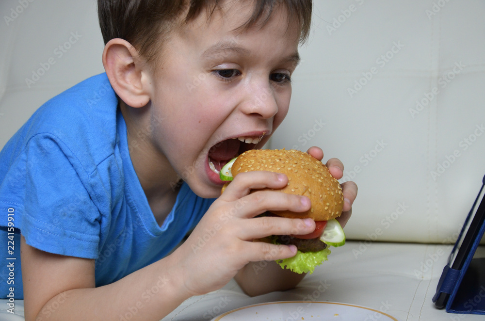 cute boy at home on the couch, eating a hamburger and looking at the ...