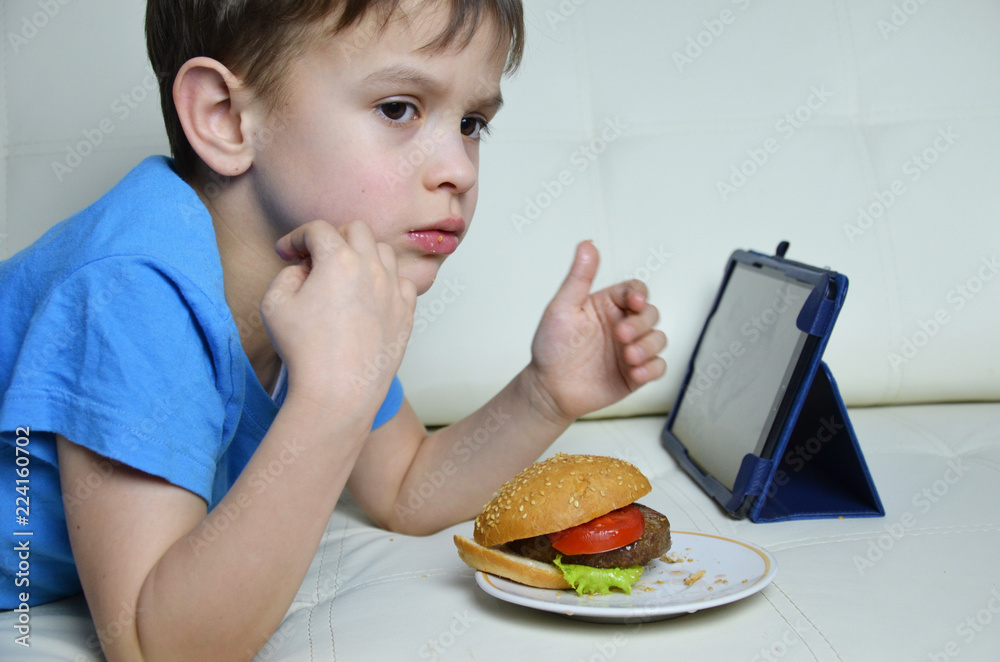 cute boy at home on the couch, eating a hamburger and looking at the ...