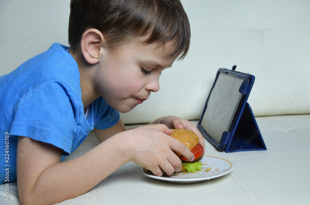 cute boy at home on the couch, eating a hamburger and looking at the ...