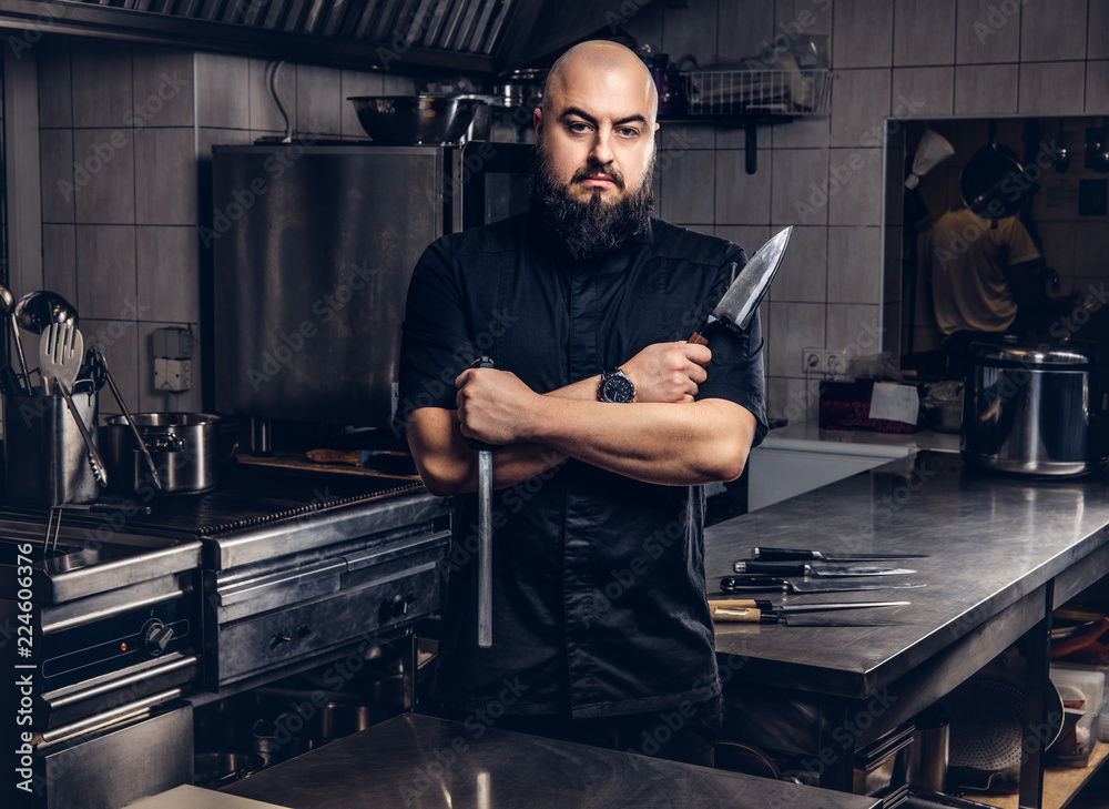Bearded cook in black uniform holds knife and standing with crossed ...