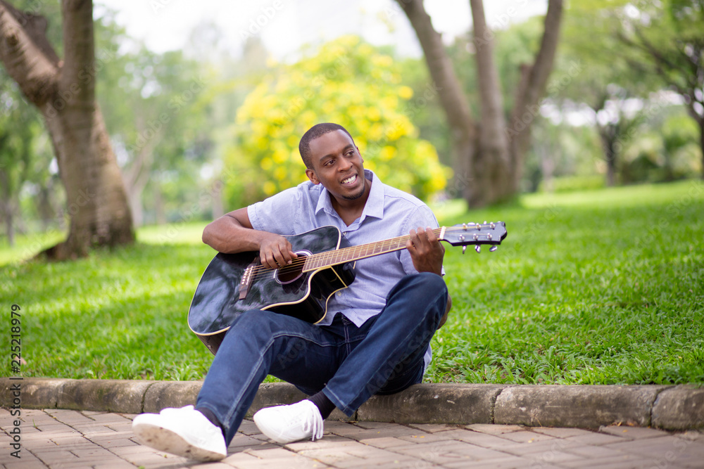 Smiling black man playing guitar and sitting on curb stone in park ...
