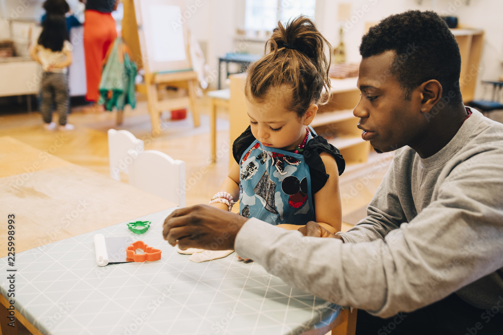 Mid adult male teacher playing with girl at table in child care ...