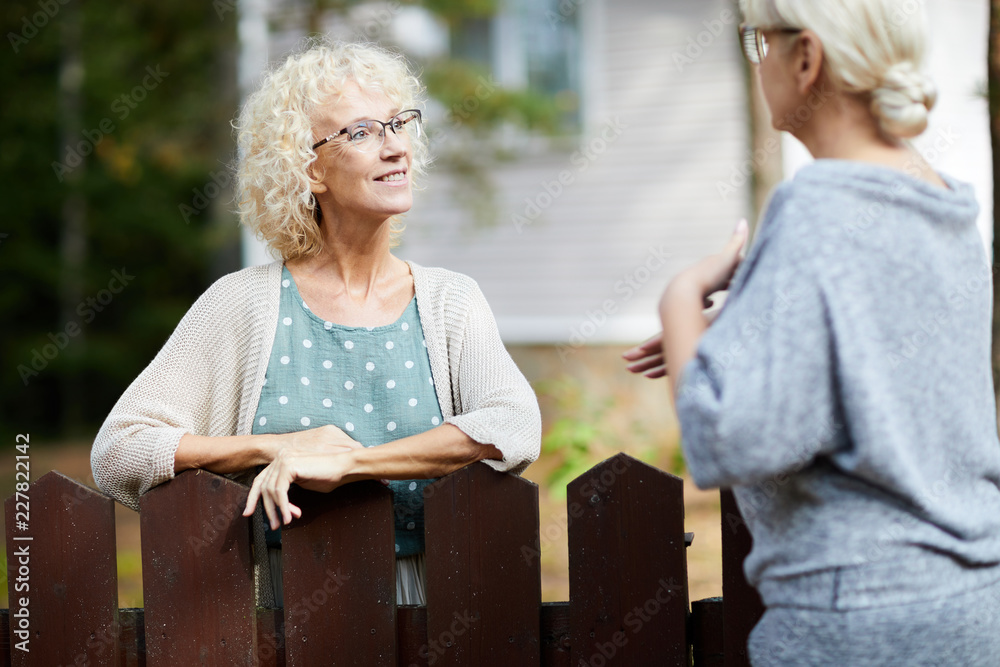 Two mature female neighbours talking through fence about everyday life ...