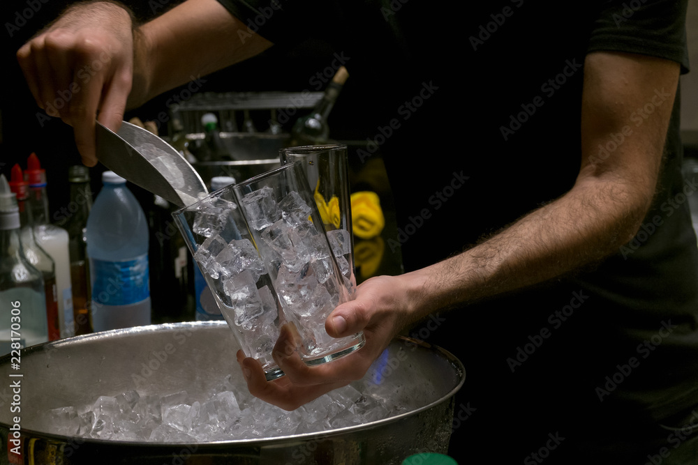 Bartender`s hand putting ice into the cocktail glass on the grey blurred background on bar ...