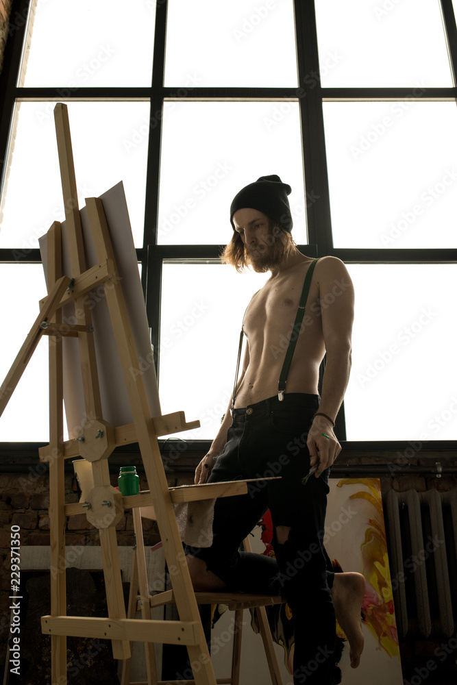 Full length portrait of shirtless contemporary artist standing by easel ...