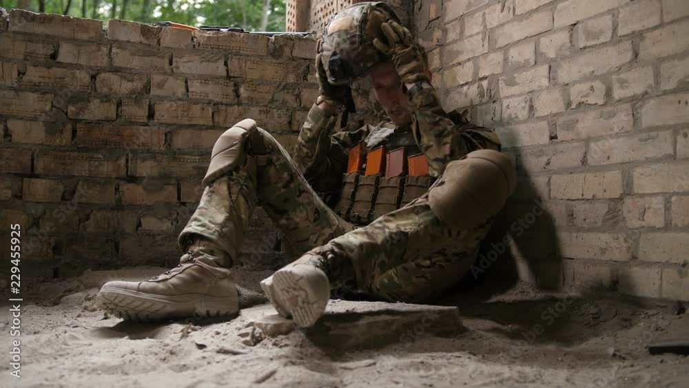 Tired after battle army soldier taking helmet off while sitting on the ground and leaning on ...