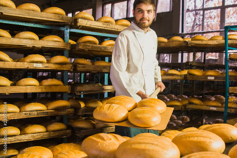 Baker. A young handsome bakery worker on the background of bread, takes ...
