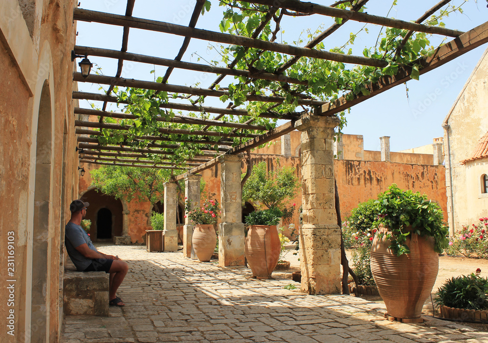 Greek courtyard - A man sits in the shade of a canopy - The yard is ...