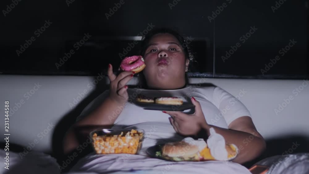 Overweight woman eats donuts and others junk foods on the bed at night ...