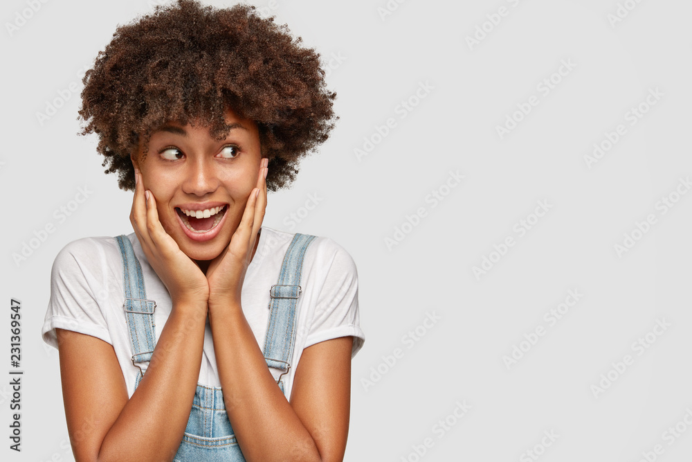 Positive young girl with dark skin, keeps hands on cheeks, looks ...