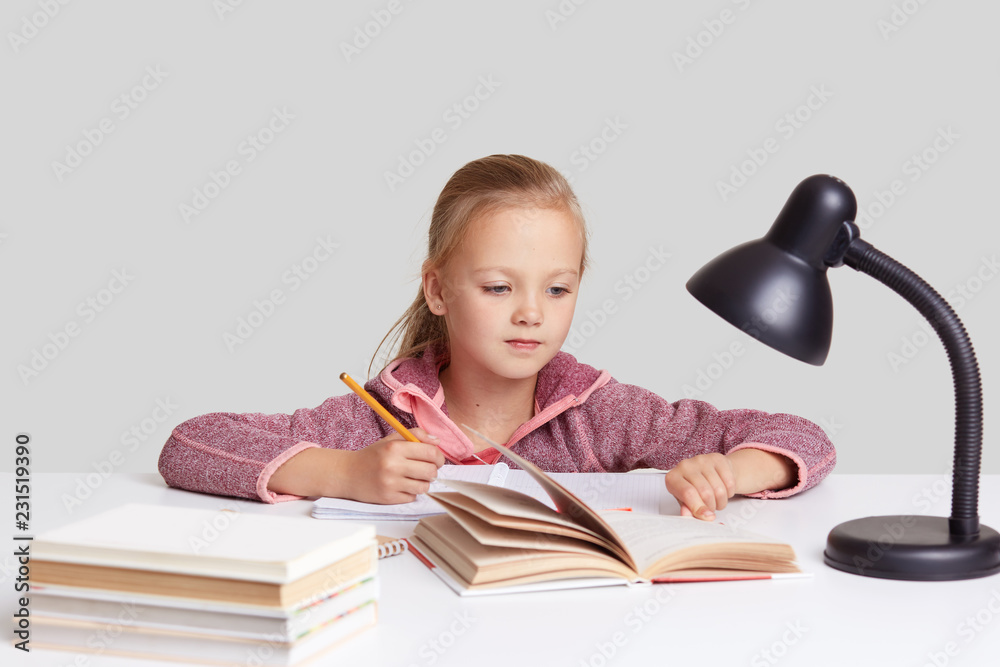 Portrait of lovely light haired schoolgirl looks attentively at book ...