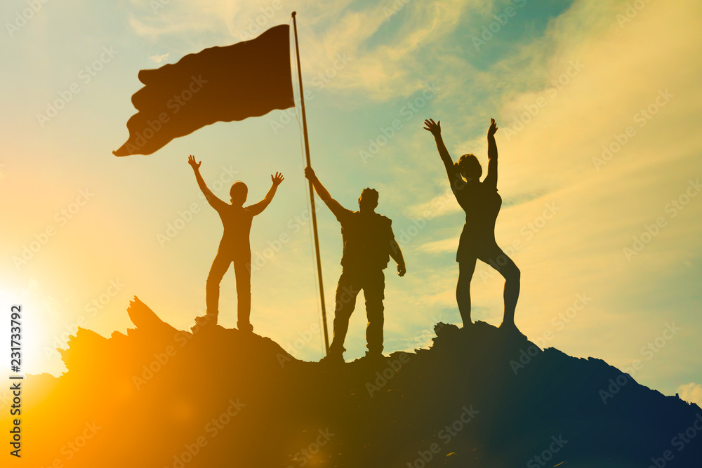 Silhouettes of happy three people on top of a mountain with the flag of ...