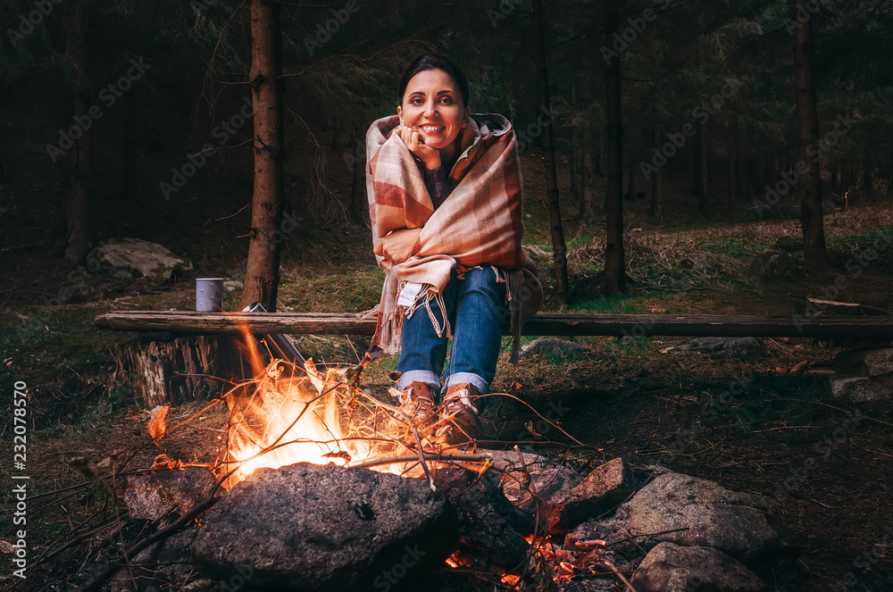 Pretty smiling young woman sits near the campfire in twilight autumn ...