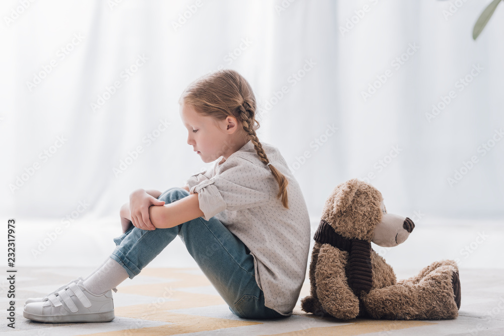 side view of sad little child sitting on floor back to back with teddy ...