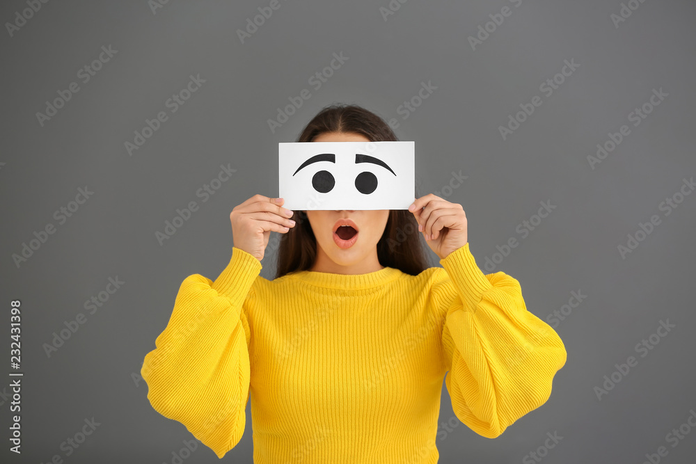 Emotional young woman hiding face behind sheet of paper with drawn eyes ...