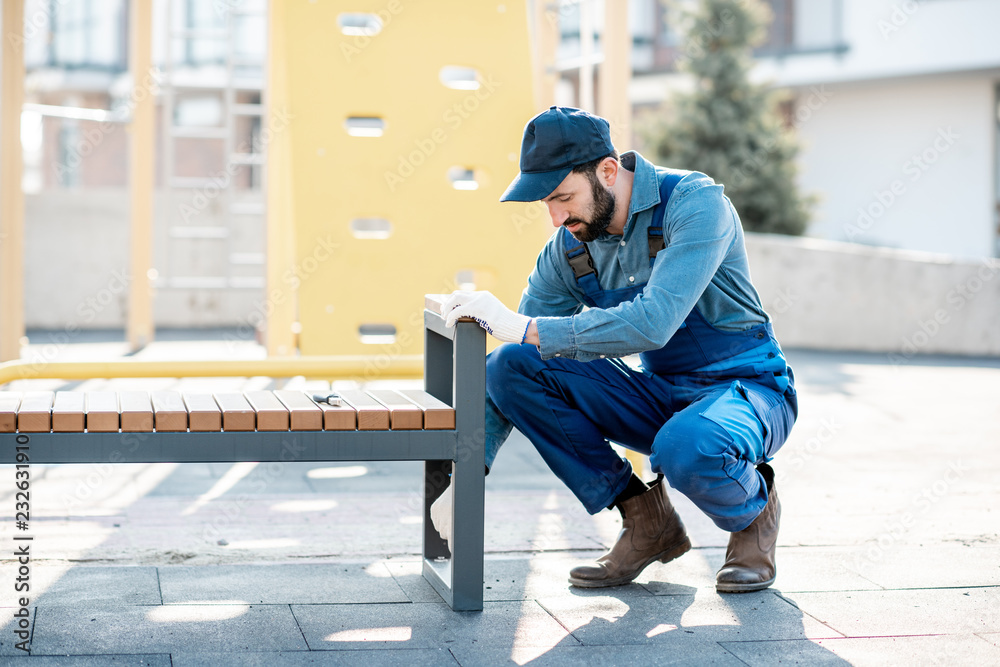 Handsome workman in uniform mounting a new bench on the playground ...