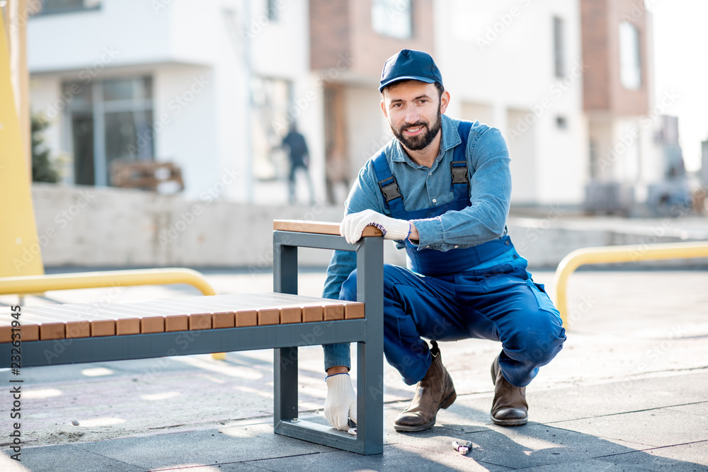 Handsome workman in uniform mounting a new bench on the playground ...