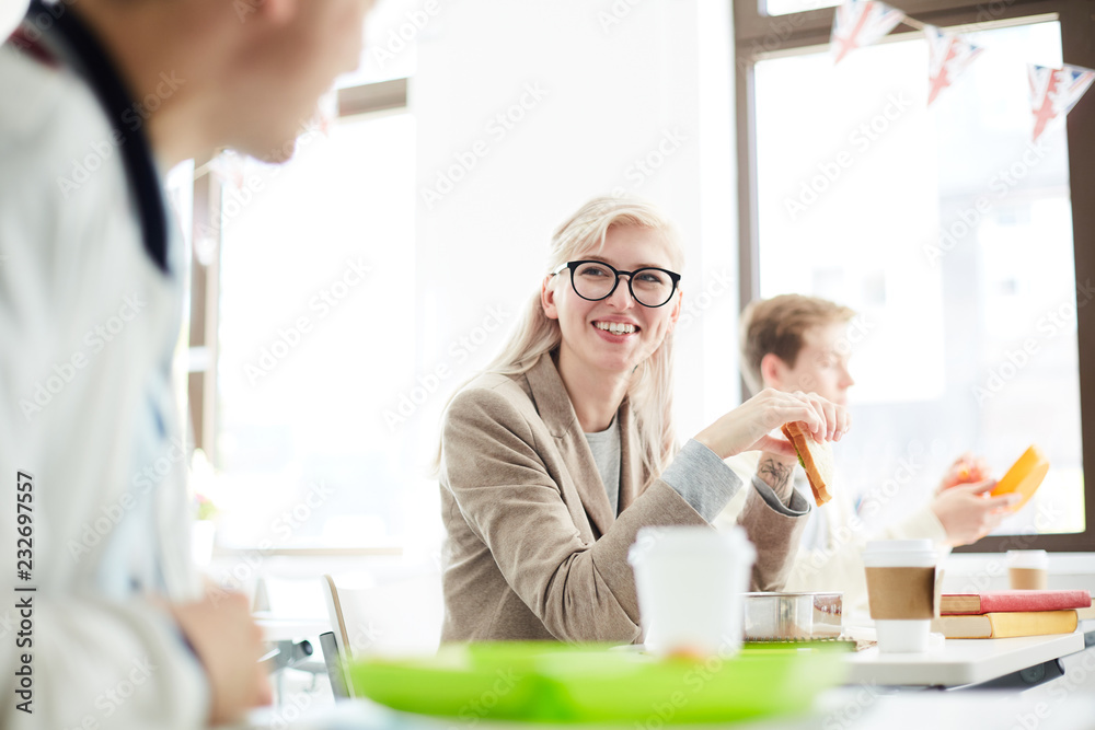 Joyful blond girl with sandwich looking at one of groupmates at lunch ...
