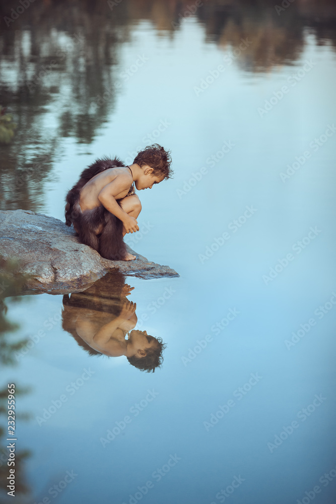 Caveman boy sitting on the rock and looking at him self in the water ...