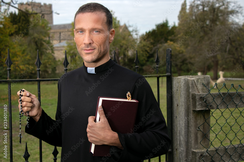 Good looking priest standing next to iron gate holding bible with field ...