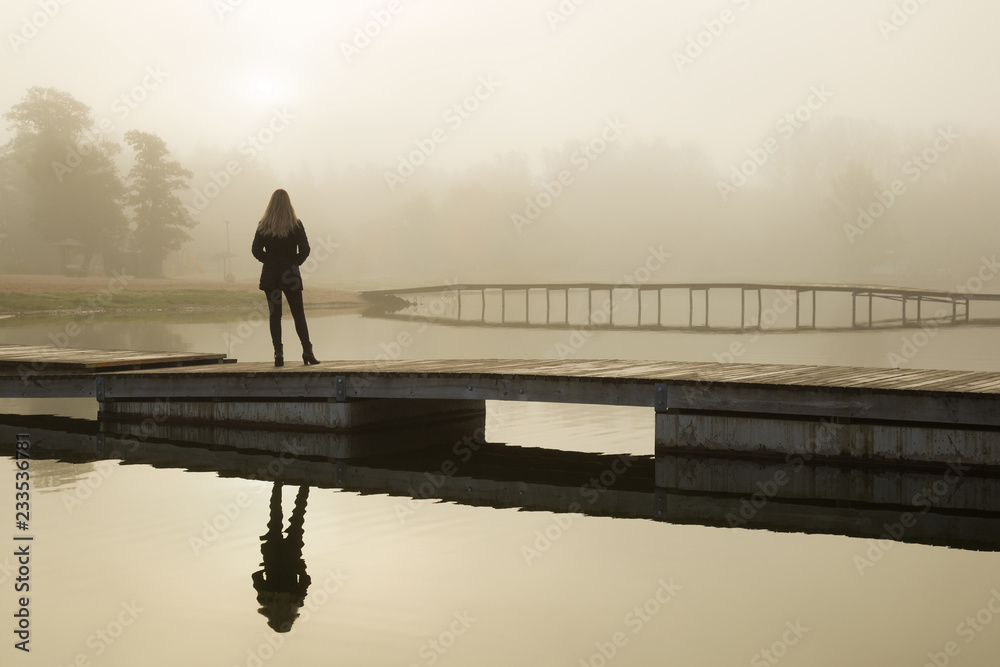 Young woman standing on lake footbridge and staring at sunrise in ...
