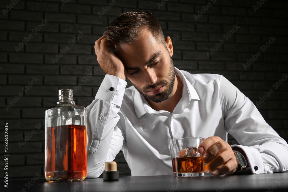 Drunk man with glass and bottle of whiskey sitting at table on dark ...