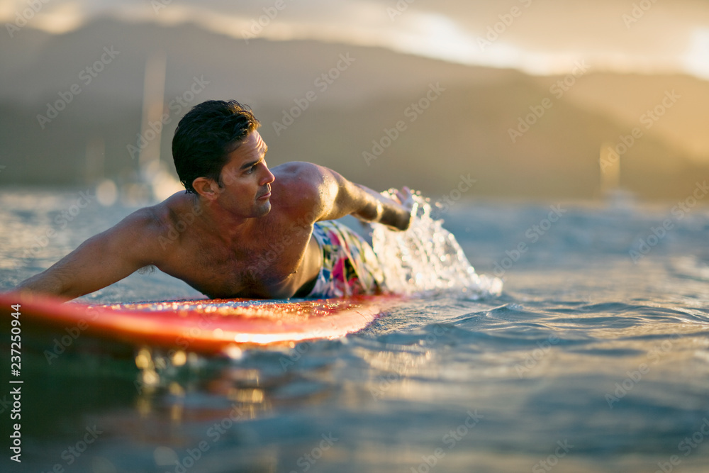 Male surfer lies on his board, looks over his shoulder and paddles in ...