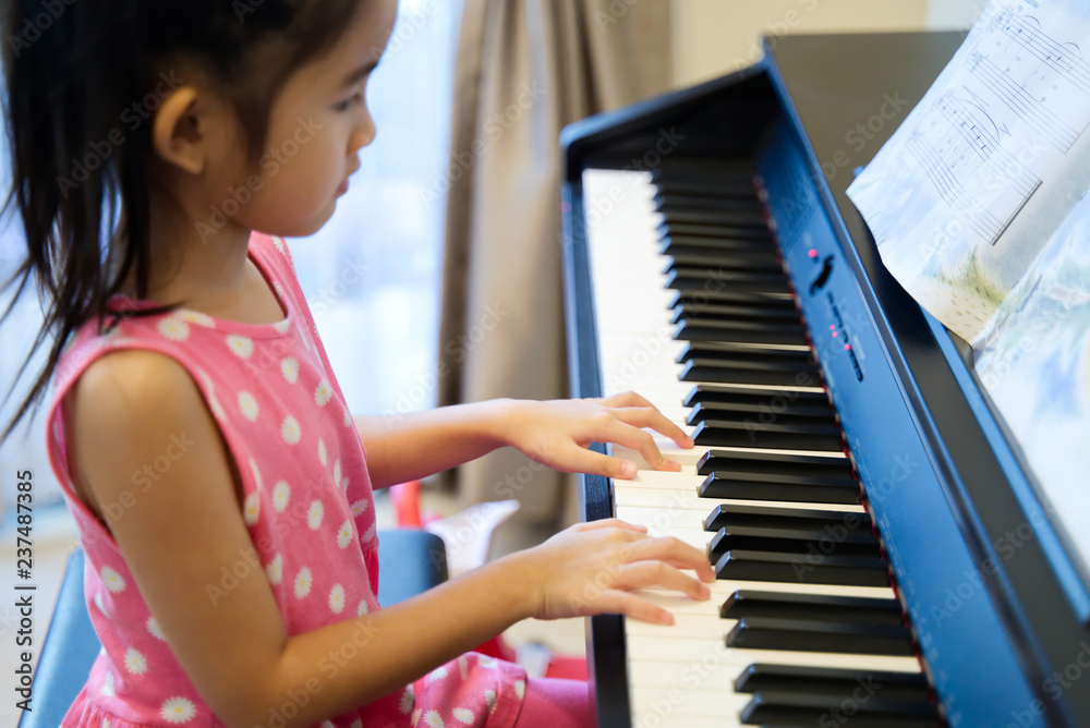 Young cute Asian kid girl playing electric piano at home. She is ...