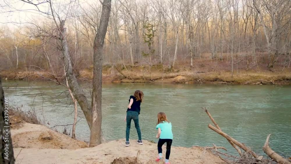 Two young girls playing by the river and throwing rocks into the water ...