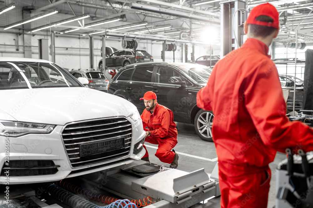 Two handsome auto mechanics in red uniform making wheel alignment with ...