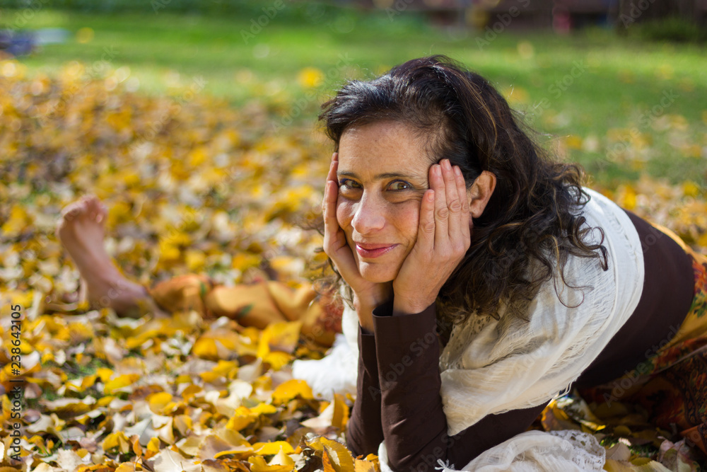 Smiling mature woman sitting on yellow and brown leaves with open legs ...