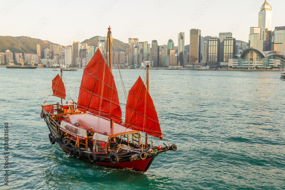 Traditional red Chinese Junk boat sailing in front of Hong Kong skyline during the day. Boat has ...