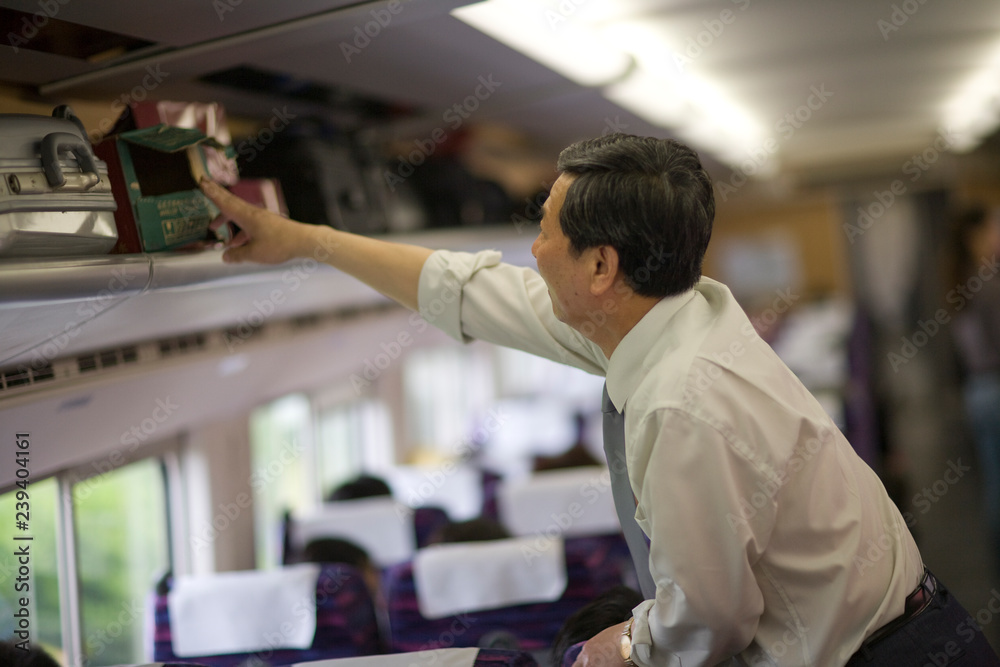 Mature adult businessman packing a bag in an overhead locker on a train ...