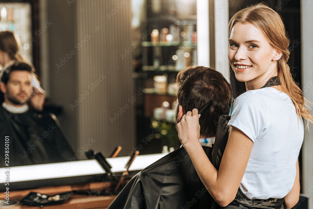 beautiful young hairstylist smiling at camera while combing hair to ... beautiful young hairstylist smiling at camera while combing hair to ...