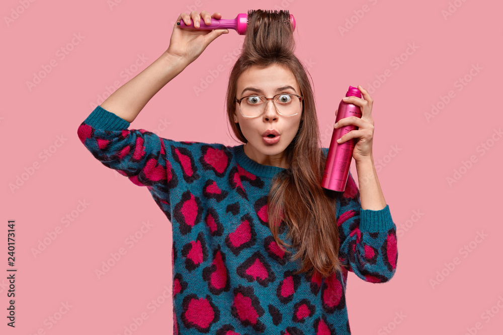 Shot of surprised woman does hair, spray fringe, uses hairbrush, wears ...