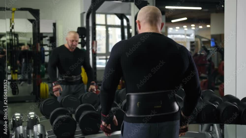 Bodybuilder takes heavy dumbbells, looks at himself in the mirror, does ...