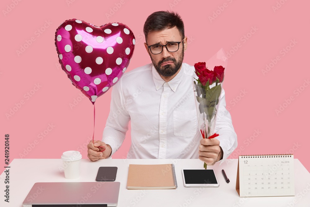 Sad bearded young male carries balloon, roses, wears glasses, sits at ...