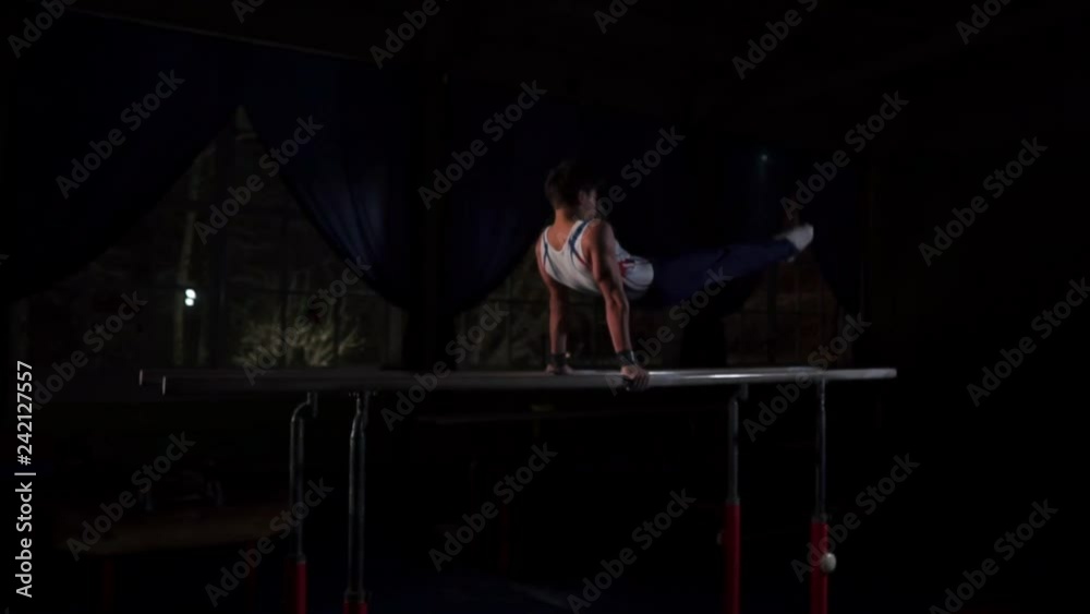 Male gymnast acrobat performs handstand on parallel bars in a dark room ...