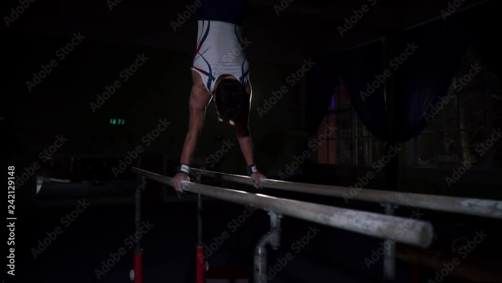 Male gymnast acrobat performs handstand on parallel bars in a dark room ...