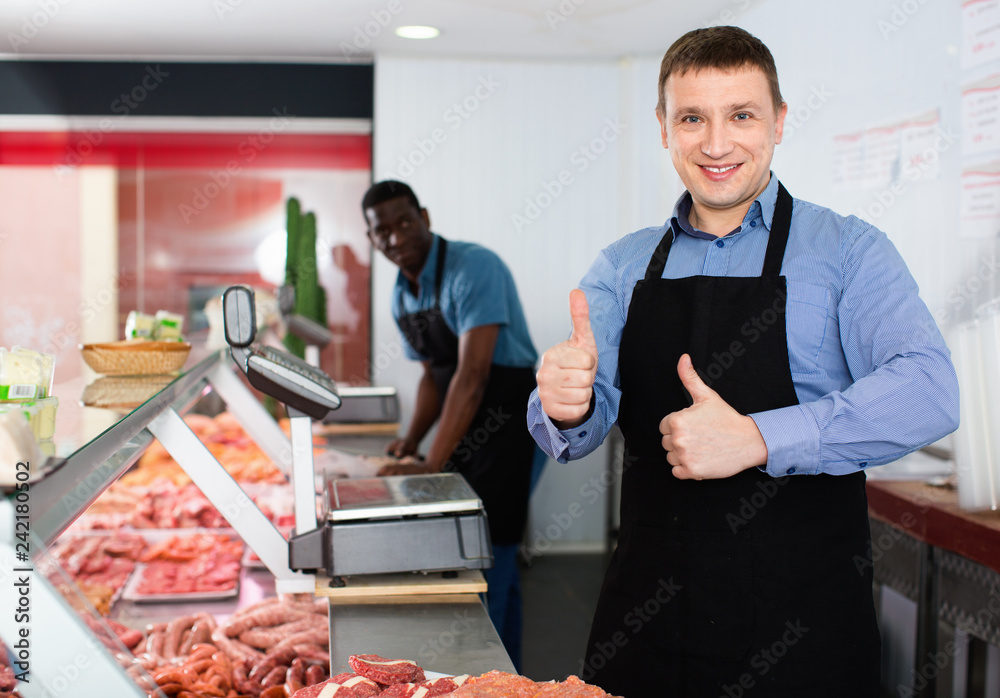Proud male owner of butcher shop standing behind counter, giving thumbs ...
