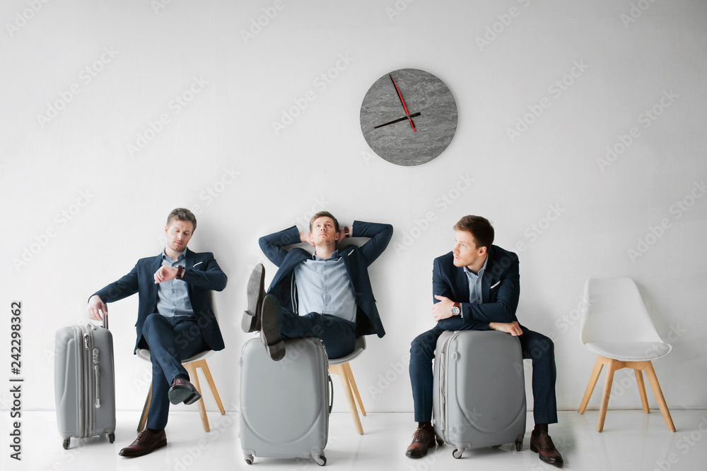 Three young men sit on white chairs and wait for flight. They look ...
