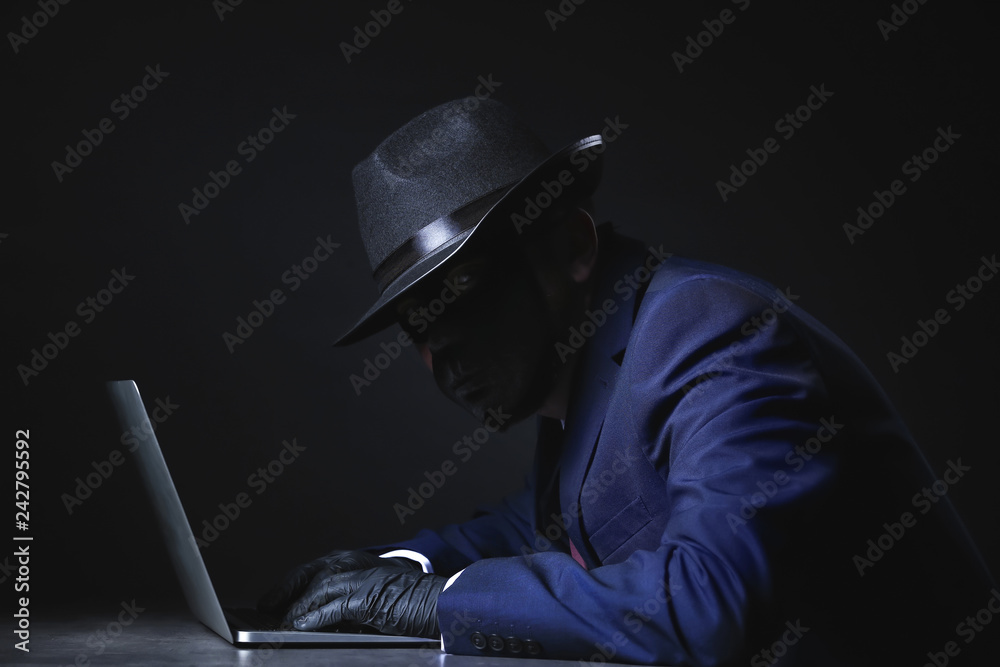 Professional hacker with laptop sitting at table on dark background ...