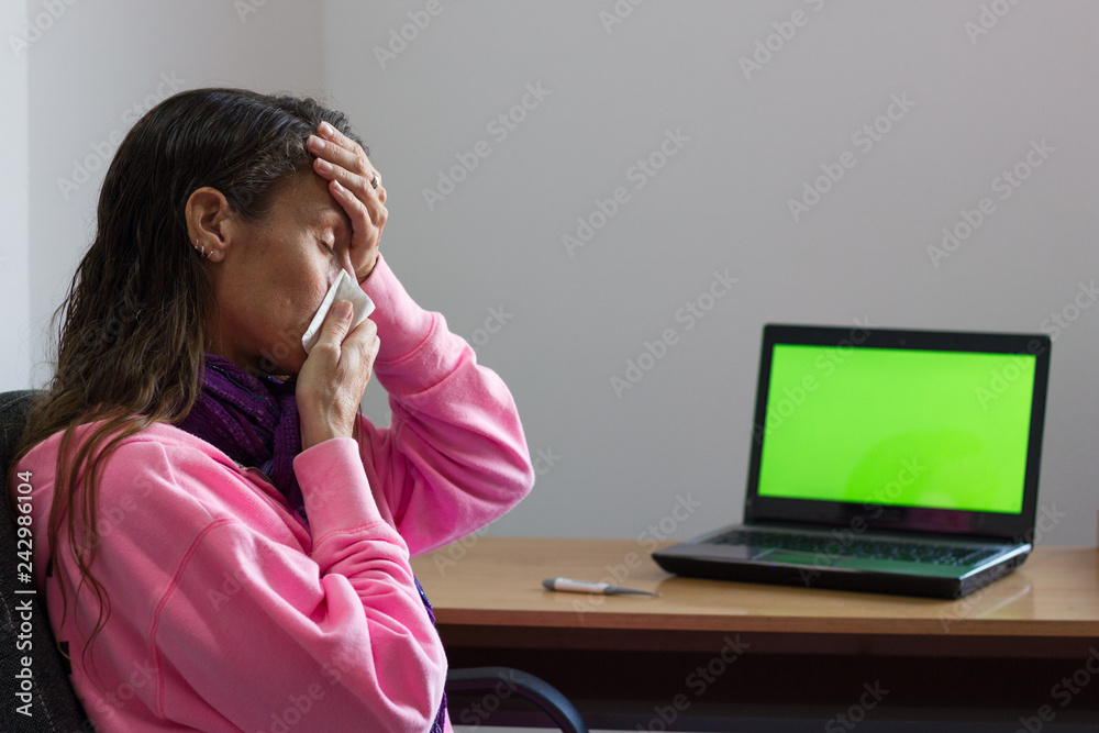 Sick lady with tissue and hand on forehead by laptop. Ill woman sitting ...