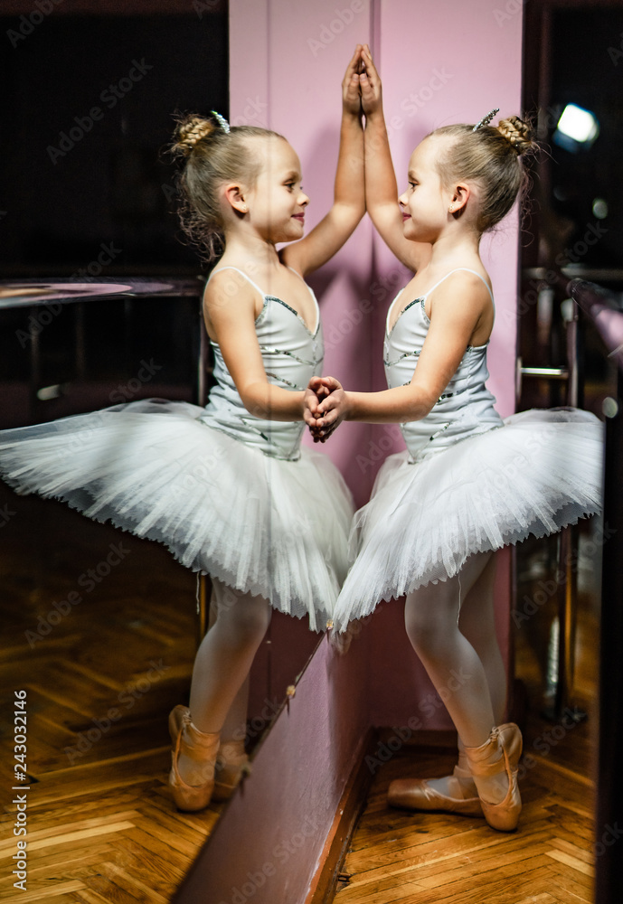 Cute little girl in a white ballet suit looking at big mirror and ...