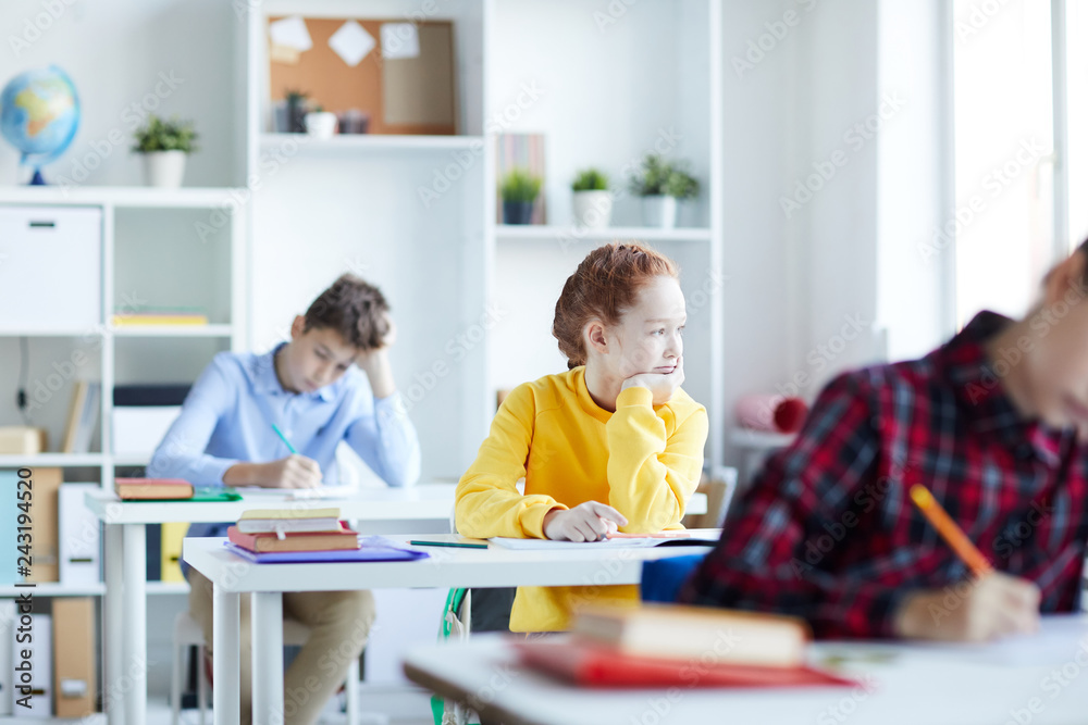 One of schoolkids sitting by desk among classmates and looking through ...