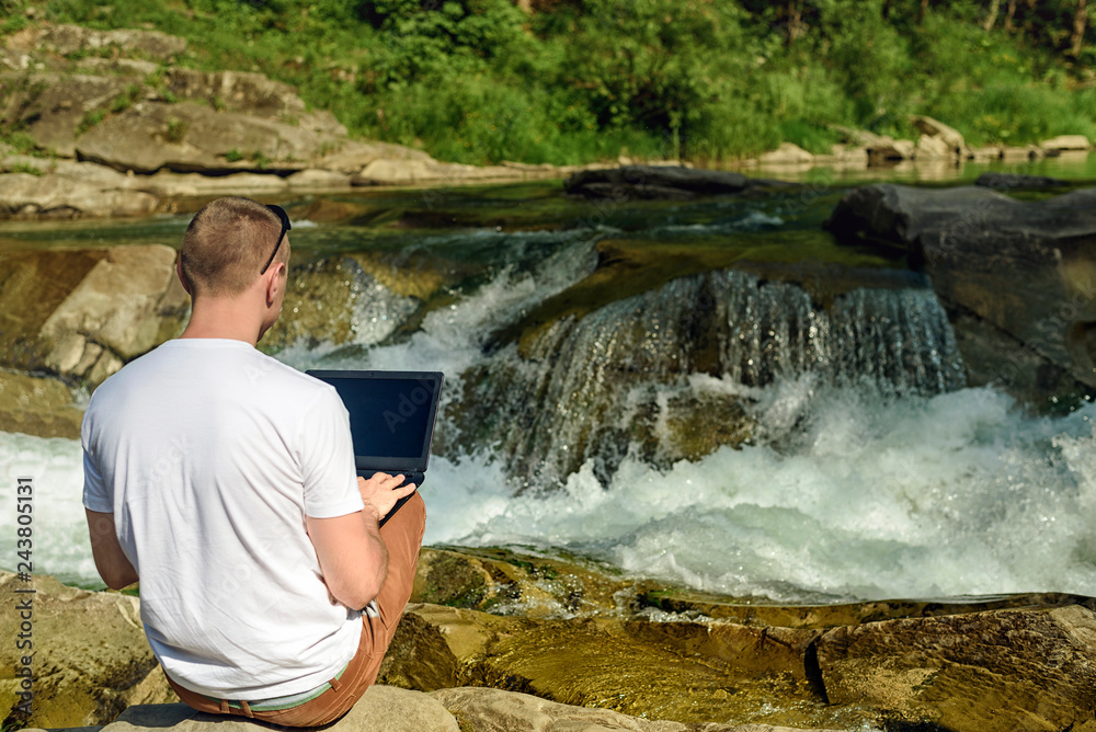 Work in nature concept. Man with notebook sitting at the bank of river ...