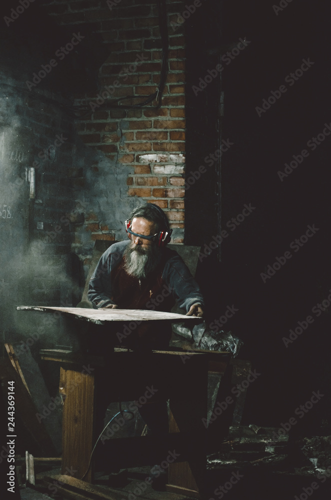 Old bearded carpenter woodworker cutting wood plate with circular saw ...