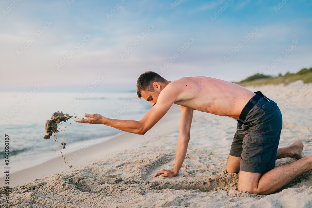 Young topless barefoot man in shorts digging hole by hands on beach ...