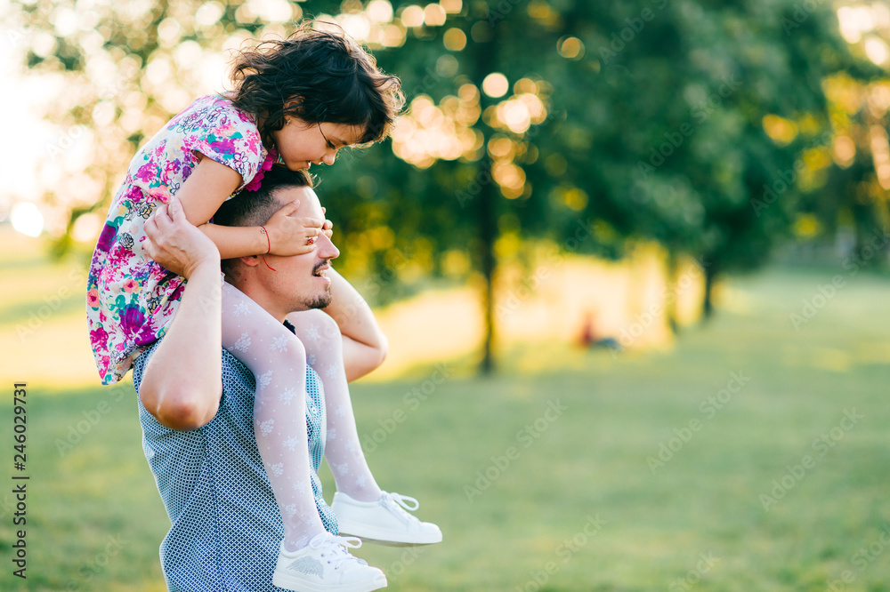 Dad carrying his daughter on shoulders in summer park at sunset. Father have fun with his little ...