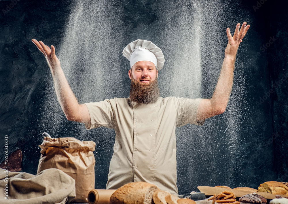 Cheerful bearded baker wearing a uniform throws up some flour standing ...
